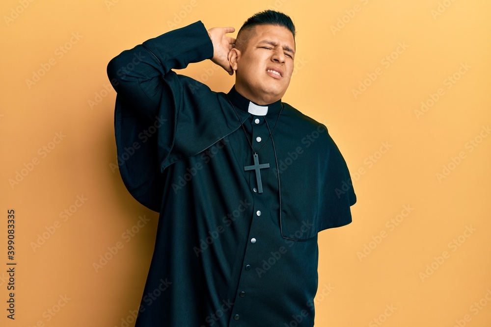 Young latin priest man standing over yellow background suffering of ...