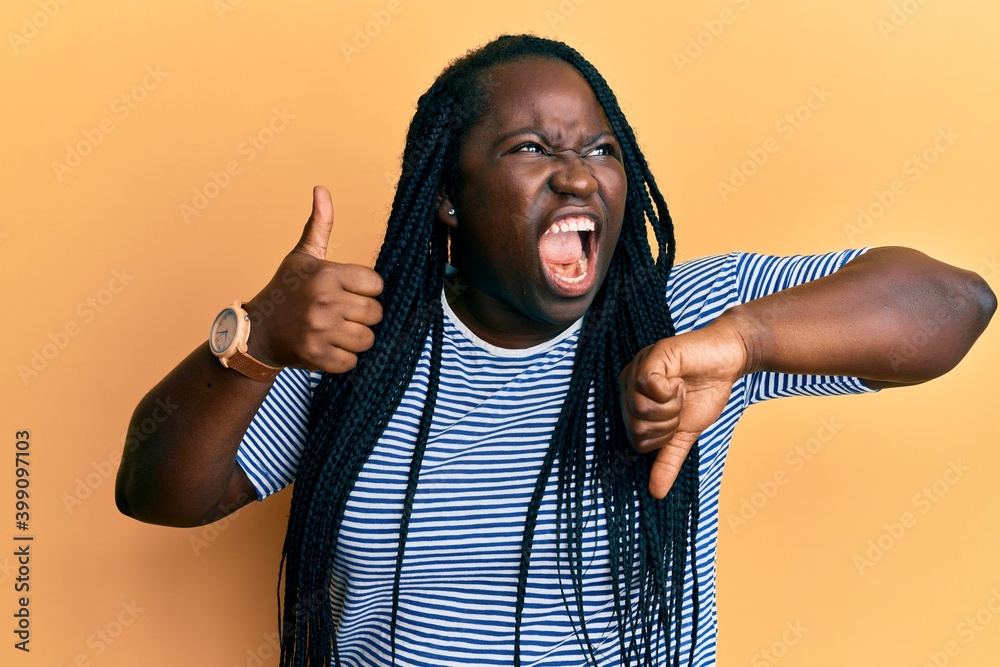 Young black woman with braids doing thumbs down and thumbs up gesture ...