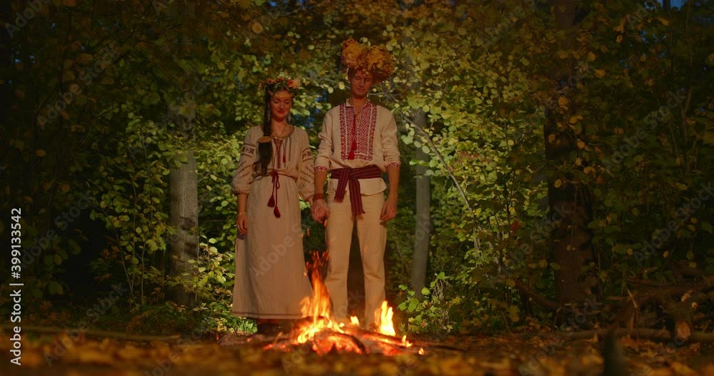 Beautiful couple in national Slavic costumes stand by the fire and hold ...