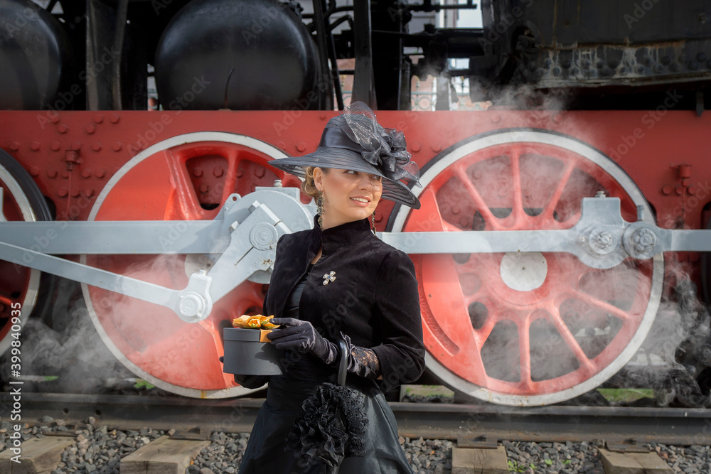 Beautiful girl in a black dress and hat near an old steam locomotive ...