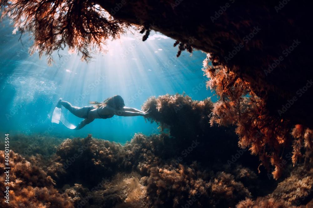 Young woman freediver girl with fins glides near rock cave with sun ...