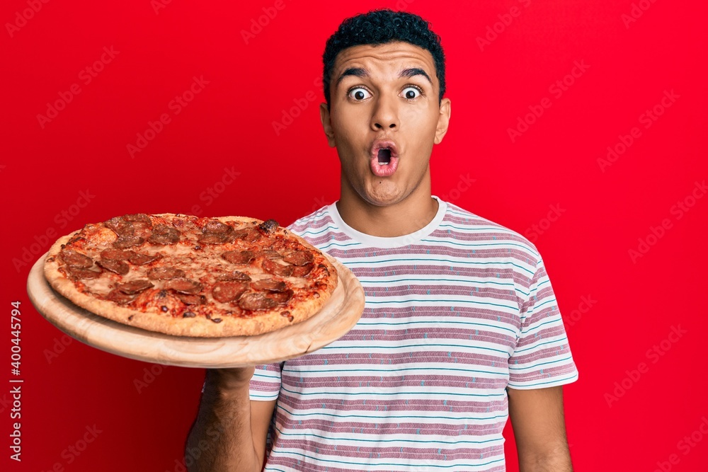 Young arab man holding italian pizza scared and amazed with open mouth ...