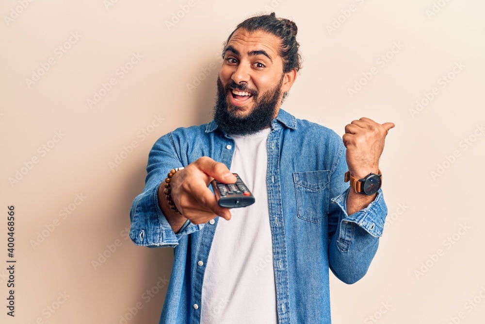 Young arab man holding television remote control pointing thumb up to ...
