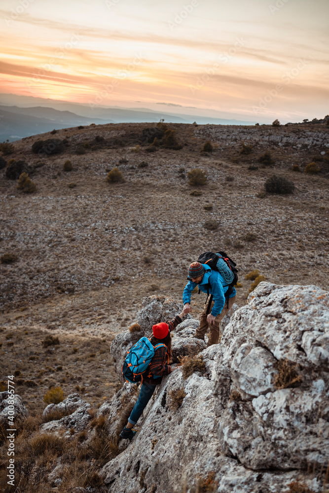 Boyfriend helping girlfriend to climb rock on mountain during sunset ...
