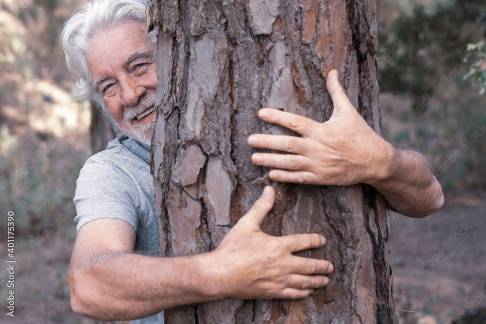 An adult man with a beard and white hair smiles as he hugs a tree trunk ...