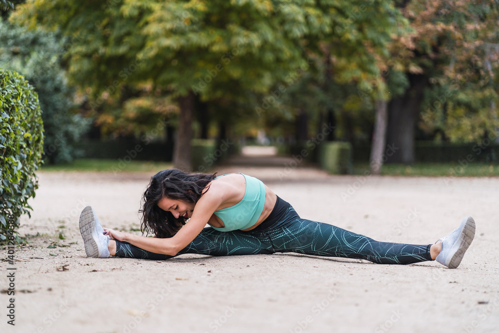 Smiling sportswoman sitting in park and stretching legs while doing ...