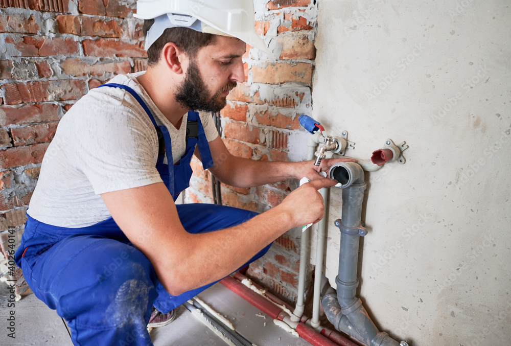 Young plumber, wearing blue uniform and white helmet working with ...