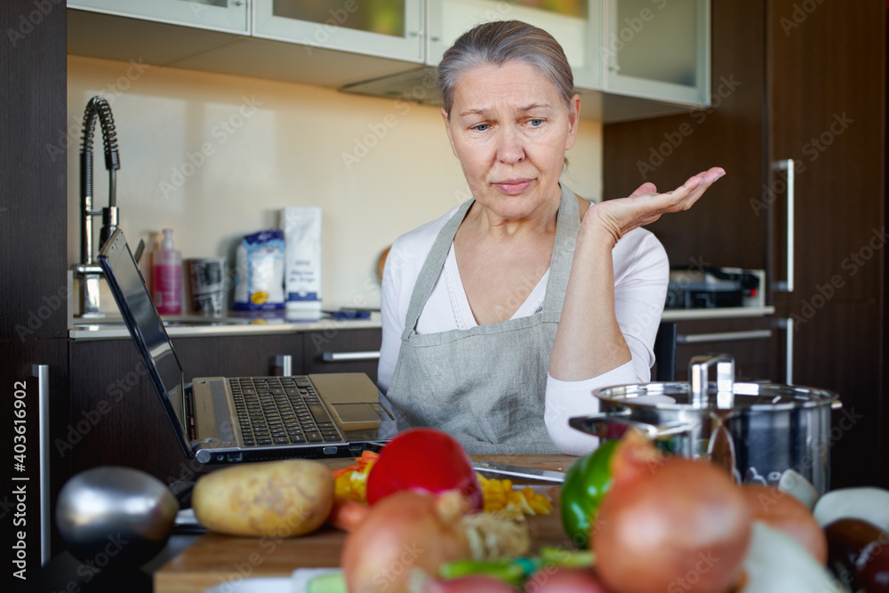 distressed mature woman in the kitchen prepares food and uses laptop ...