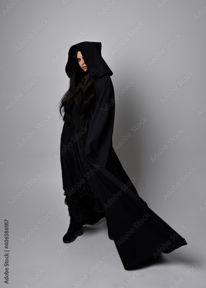 Full length portrait of pretty black haired woman wearing long dark gown and a cloak. Standing ...