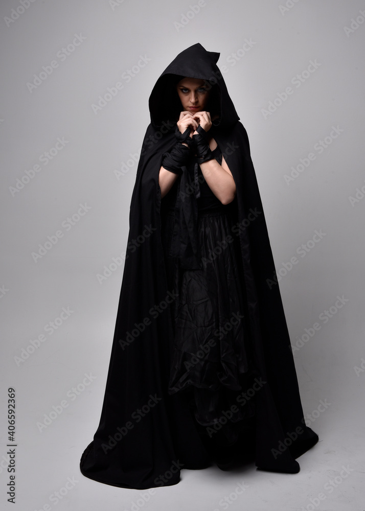 Full length portrait of pretty black haired woman wearing long dark gown and a cloak. Standing ...