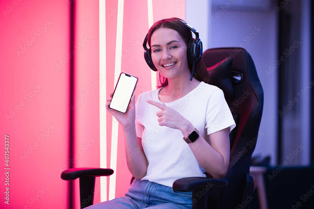 Smiling gamer girl wearing headset showing smart phone and pointing at ...