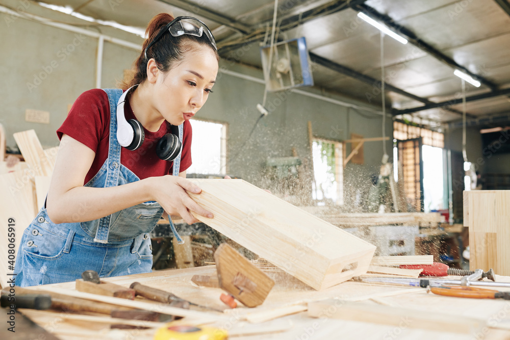 Young female Asian carpenter blowing off wooden dust from drawer she ...