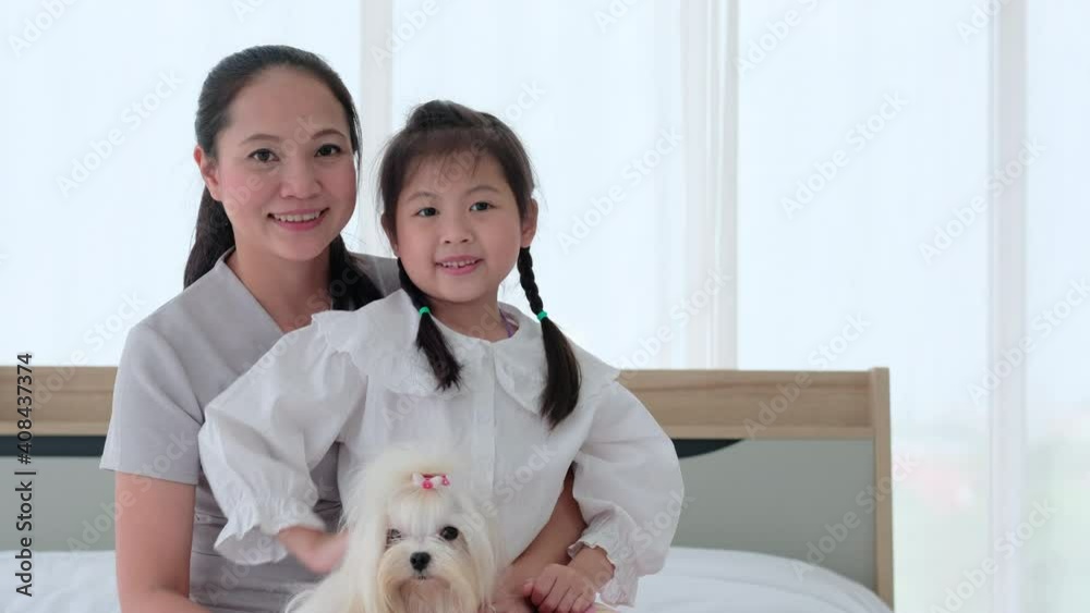 Mother and daughter sit on bed with holding dog and look at camera ...
