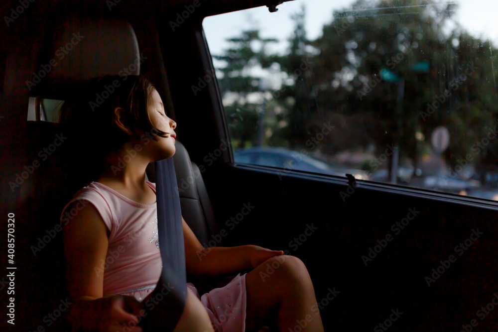 A beautiful little girl wearing seat belt sleeps in a car at twilight ...