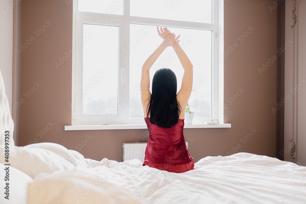 back view, Beautiful brunette in red pajama is waking up in the morning ...