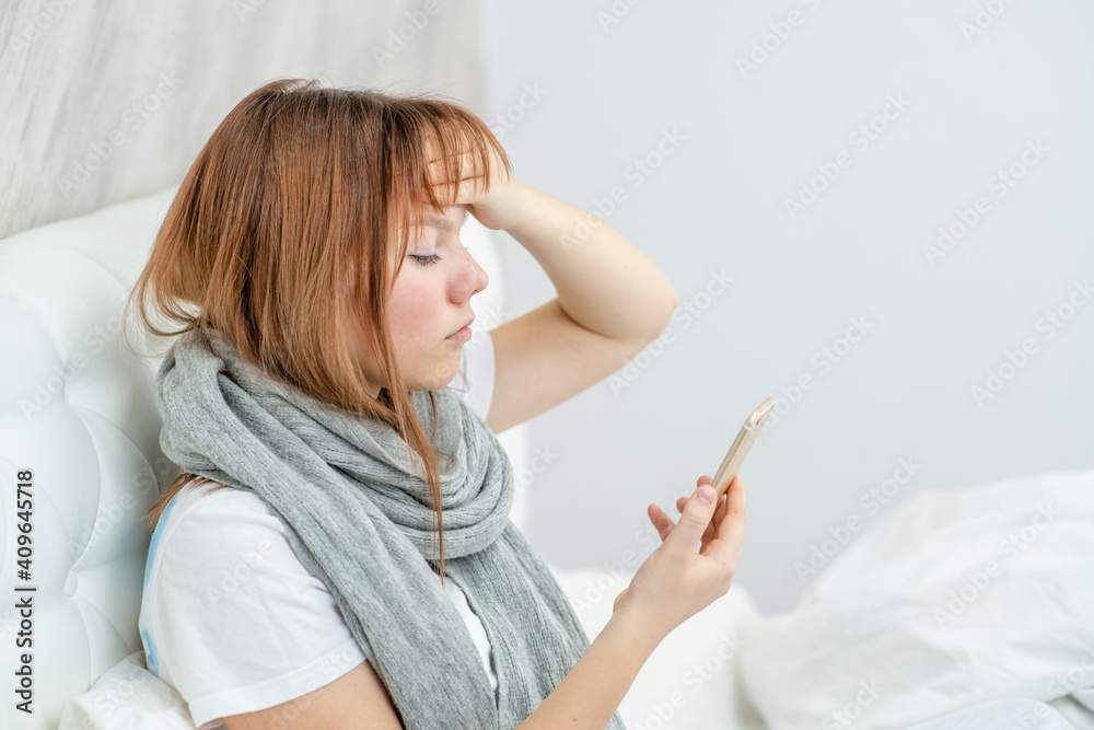 Sick girl sits at home on a bed, touch her forehead checking ...