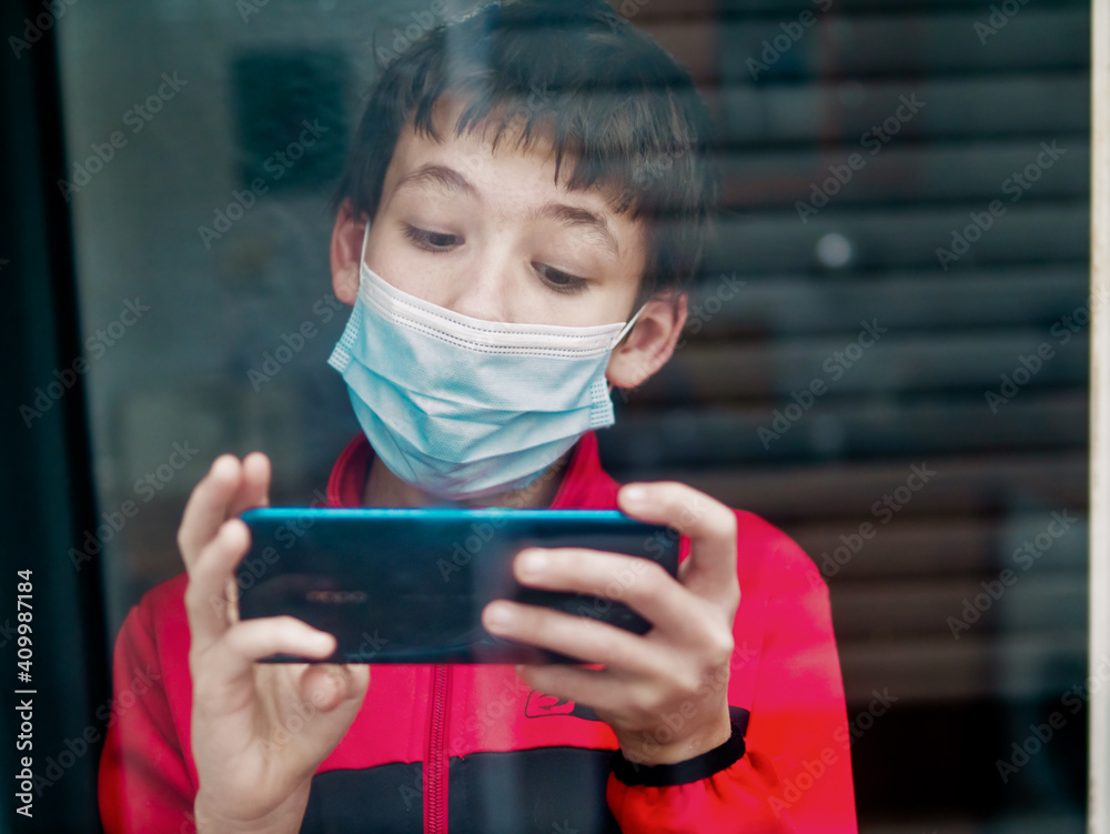 Teenage boy with medical mask at home behind a window using his smart ...