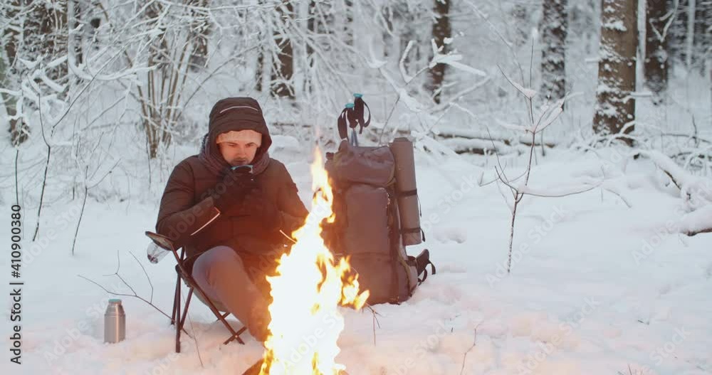 Caucasian man sitting by campfire in snowy woods and drinks hot tea ...
