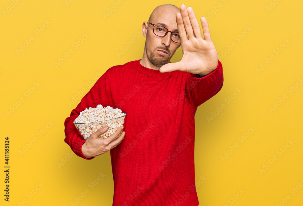 Bald man with beard eating popcorn with open hand doing stop sign with ...