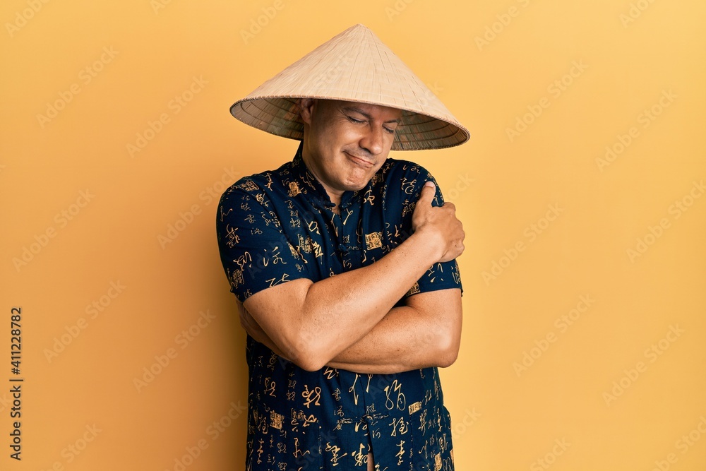Middle age bald man wearing traditional asian straw hat hugging oneself ...