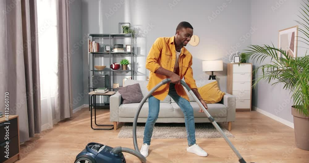 African American joyful happy handsome young guy vacuuming living room ...