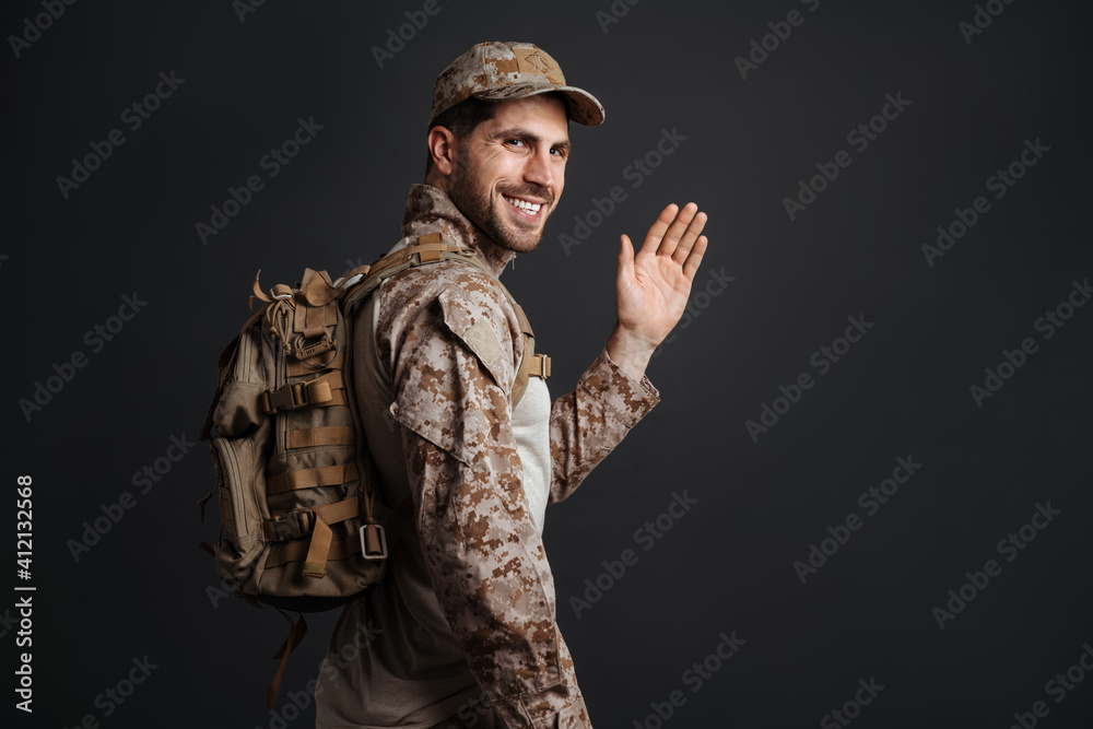 Smiling masculine military man waving hand while posing with backpack ...