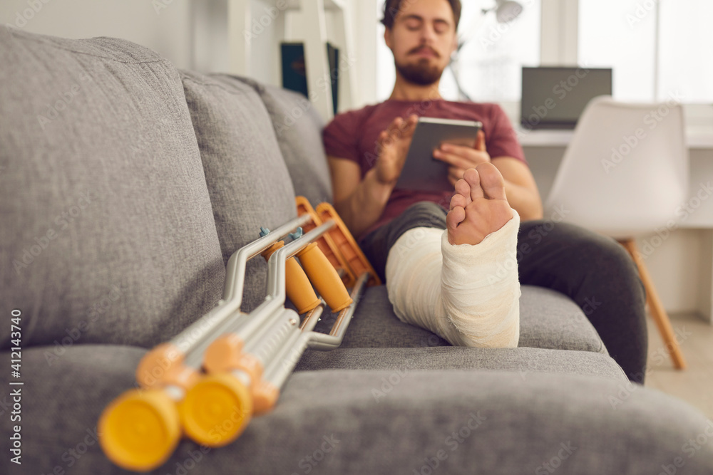 Young man with broken leg in plaster cast sitting on sofa and using ...