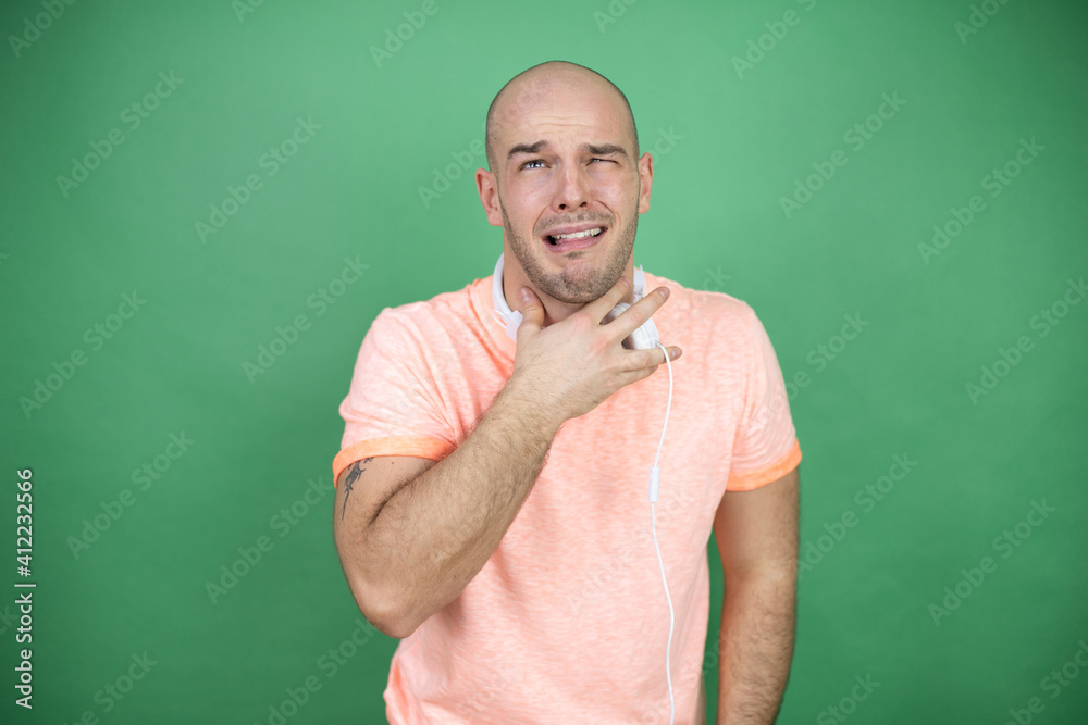 Young bald man using headphones over green background touching painful ...