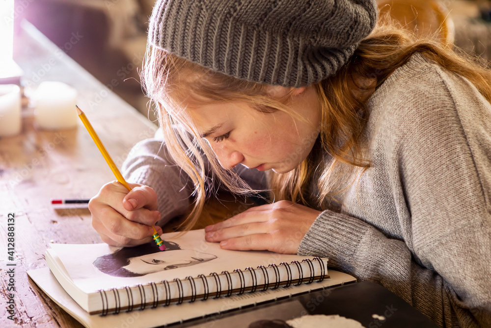 Teenage girl in a woolly hat drawing with a pencil on a sketchpad ...