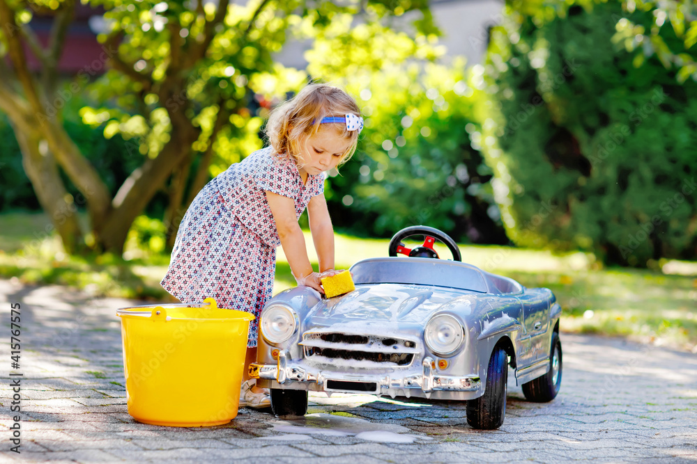 Cute gorgeous toddler girl washing big old toy car in summer garden ...