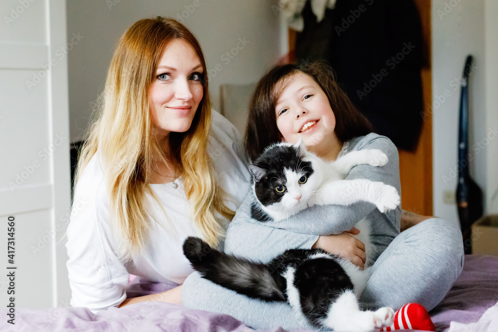 Lovely young mother and cute school kid girl cuddling together with cat ...