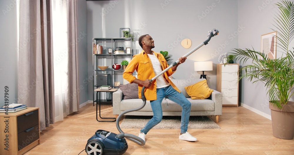 African American joyful happy handsome young guy vacuuming living room ...