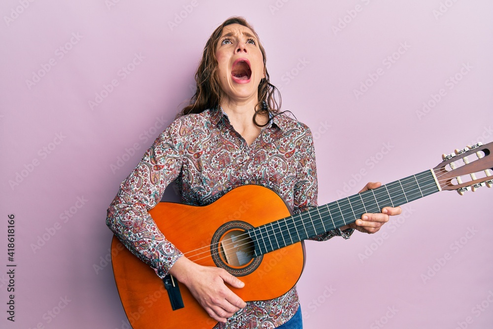 Young blonde woman playing classical guitar angry and mad screaming ...