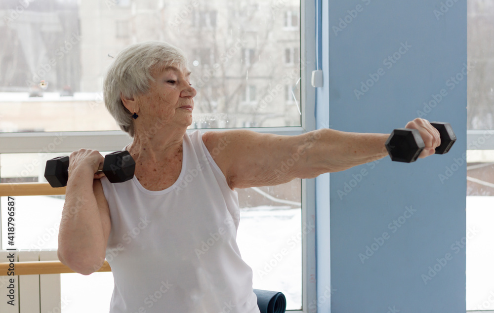 An older athletic woman does a dumbbell exercise in a light gym. Active ...