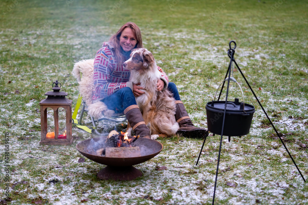 An Australian Shepherd sits by the campfire with his owner in the afternoon. Snow on grass ...