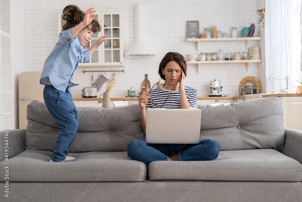 Irritated Mother Sitting On Couch Working On Laptop At Home Distracted
