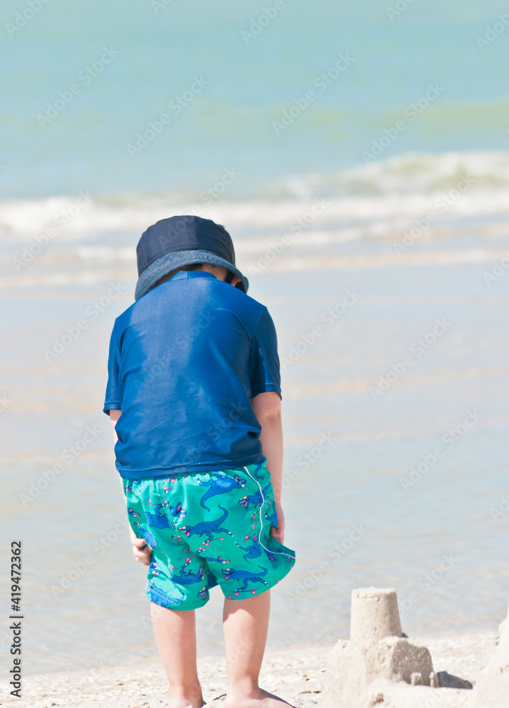 back view of a young boy dressed in blue, hat, shirt and bathing suit ...