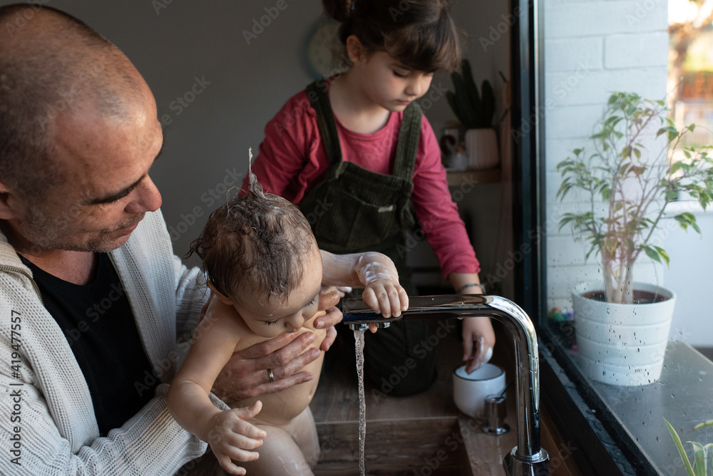 Little girl washing cute baby in arms of father during bathing in sink ...