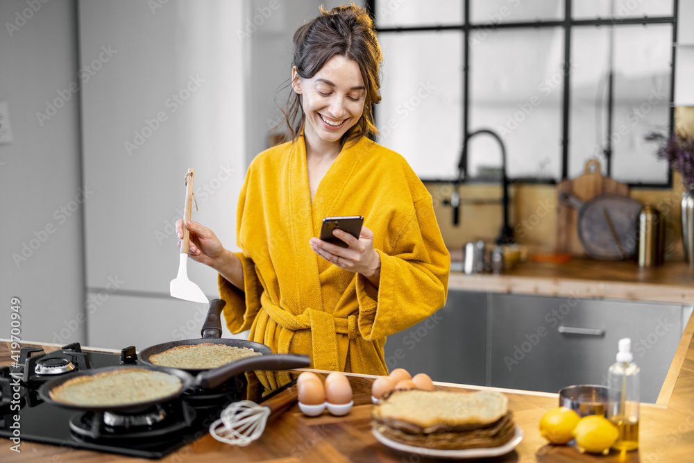 Happy woman dressed in bathrobe cooks pancakes for breakfast, looks on ...