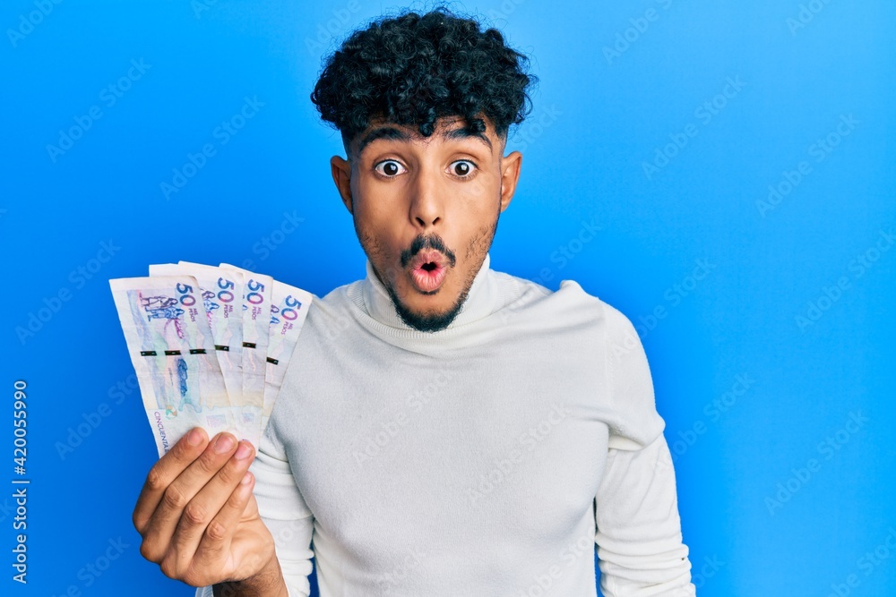 Young arab handsome man holding 50 colombian pesos banknotes scared and ...