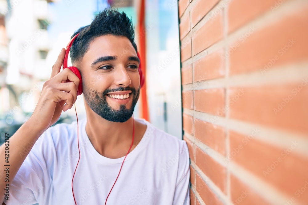 Young arab man smiling happy using headphones leaning on the wall at ...