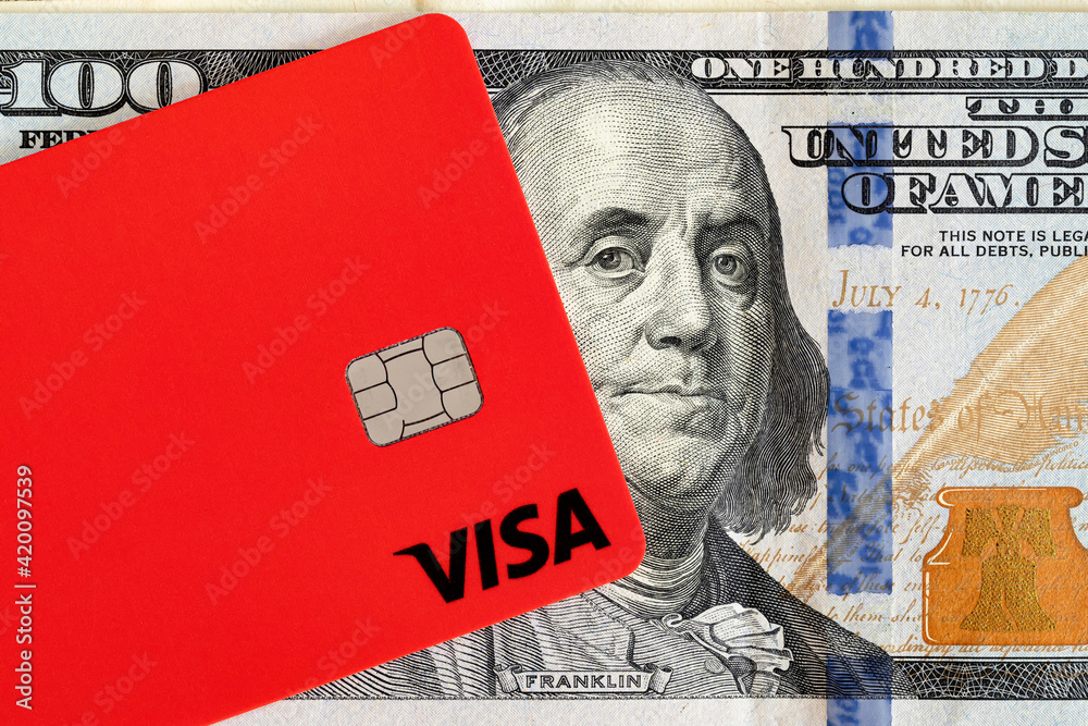 Red bank card with VISA logo on the 100 US dollar banknote. VISA is an ...