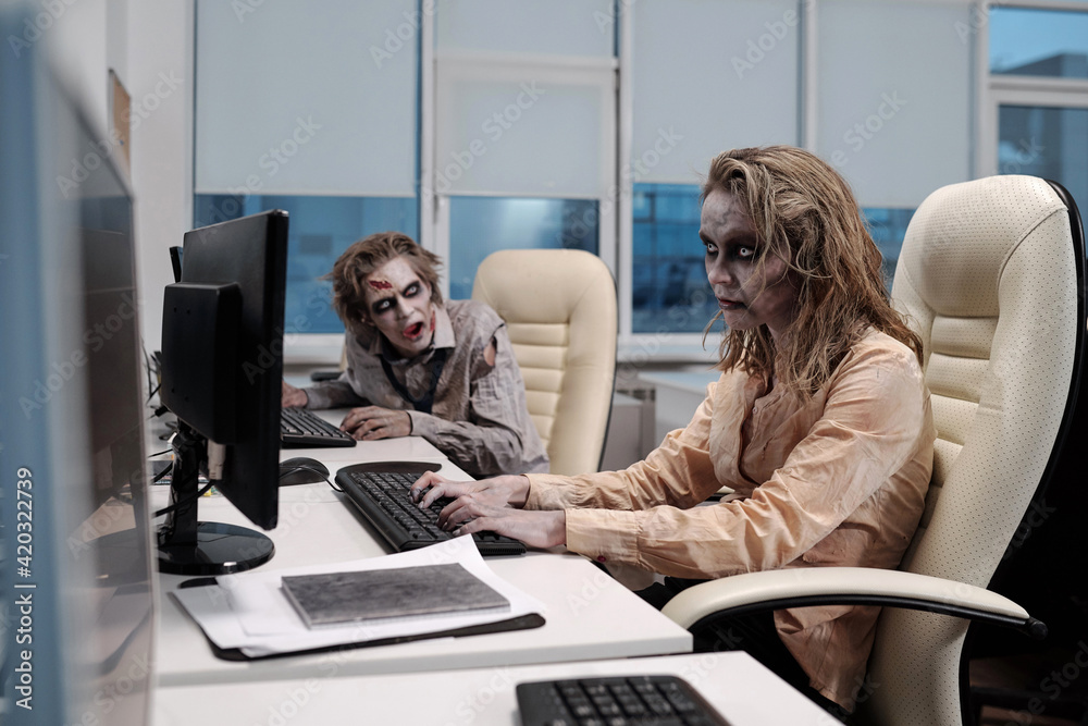 Young spooky zombie businesswoman sitting by desk in front of computer ...