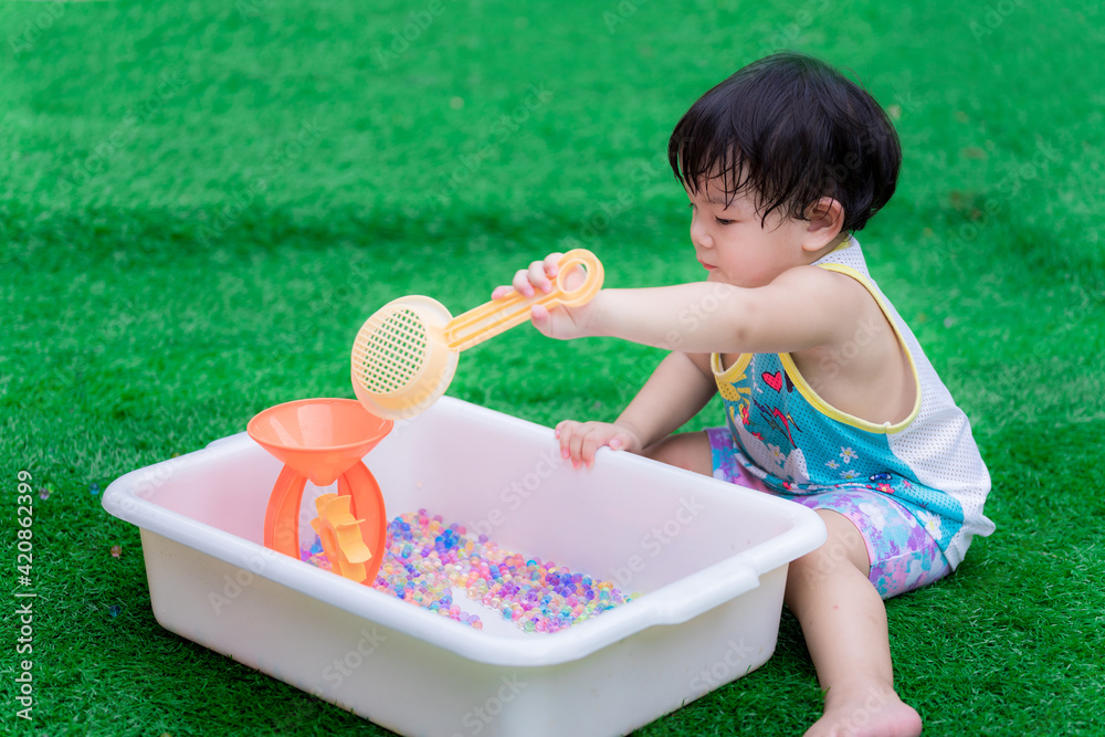 Asian preschool baby uses an orange toy sieve to scoop rainbow beads ...