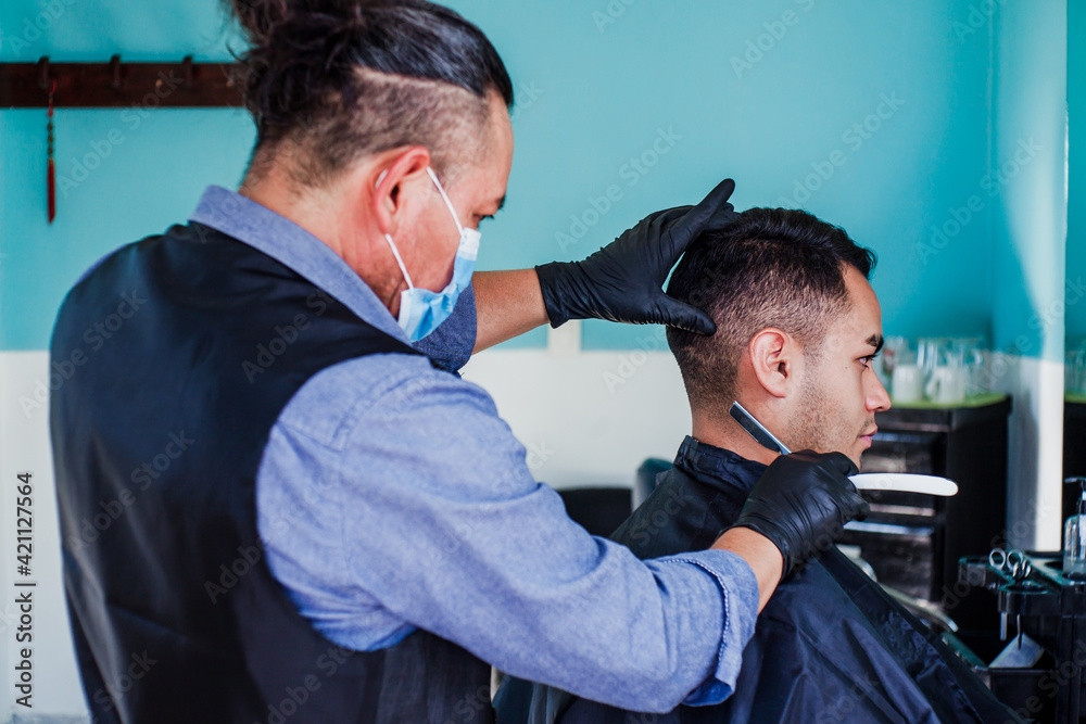 hispanic man stylist with facemask cutting hair to a client in a barber ...