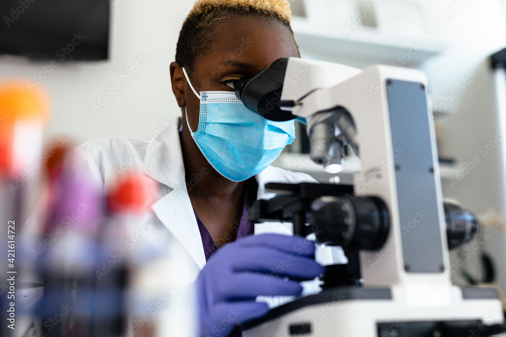 Black medical technologist in mask in laboratory looking through ...