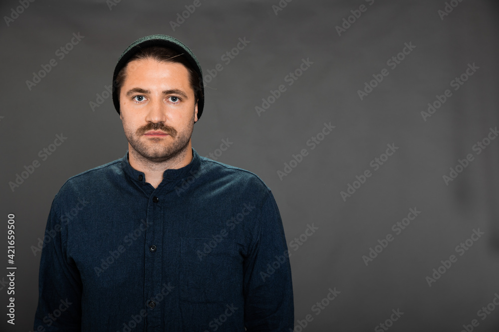 Handsome unshaven wimp wobbly guy in knitted cap posing on gray studio ...