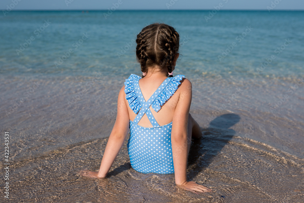 Rear view of cute girl in blue swimsuit sitting on beach. Child by the ...
