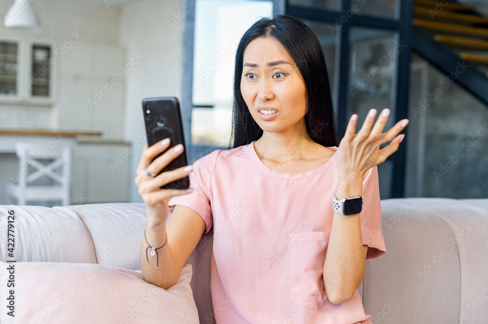 Confused asian woman holding smartphone sitting on sofa at home, stares ...