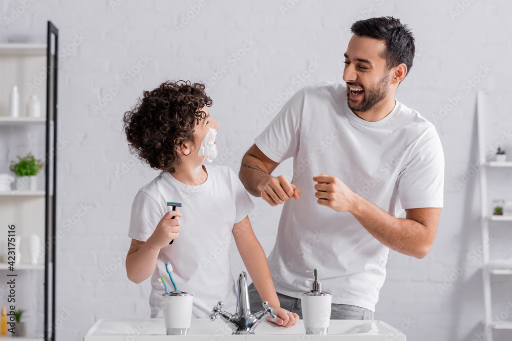 Smiling arabian father standing near son in shaving foam holding razor ...